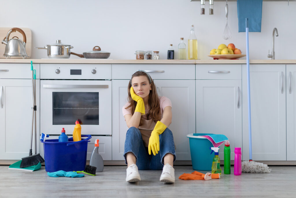 Exhausted young housewife sitting on floor at kitchen, surrounded by ...